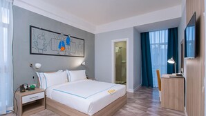Quarto de hotel com cama, mesa de cabeceira, abajur e escrivaninha com cadeira.