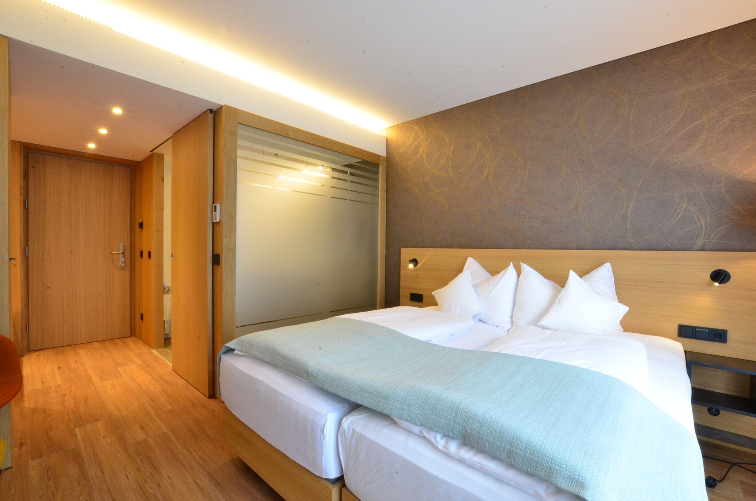 Quarto de hotel moderno com uma cama grande, piso de madeira, armário embutido e cabeceira fixada na parede.