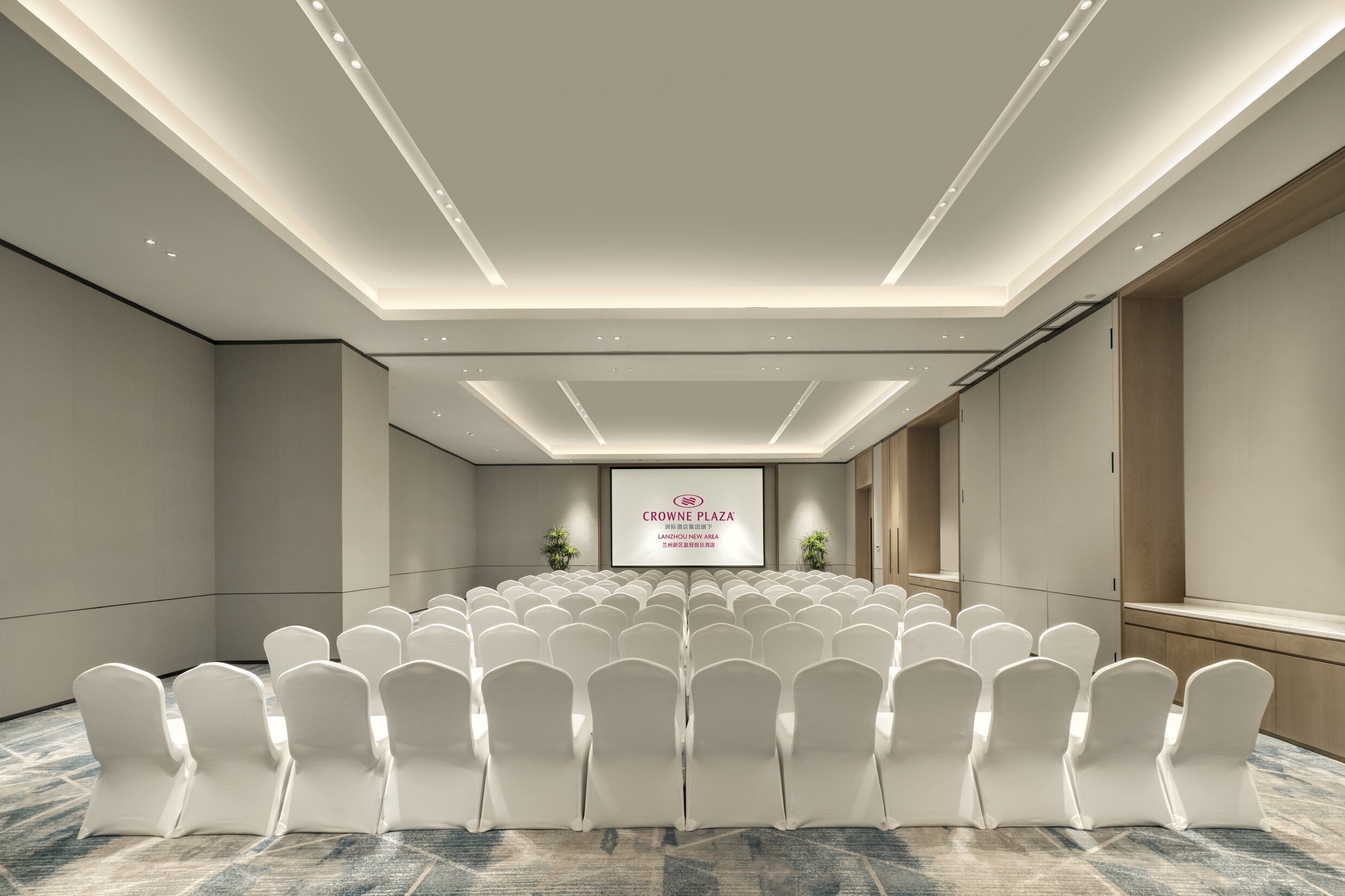 Sala de conferências com fileiras de cadeiras brancas, um púlpito e uma tela exibindo "Crowne Plaza Landhuis New Area".