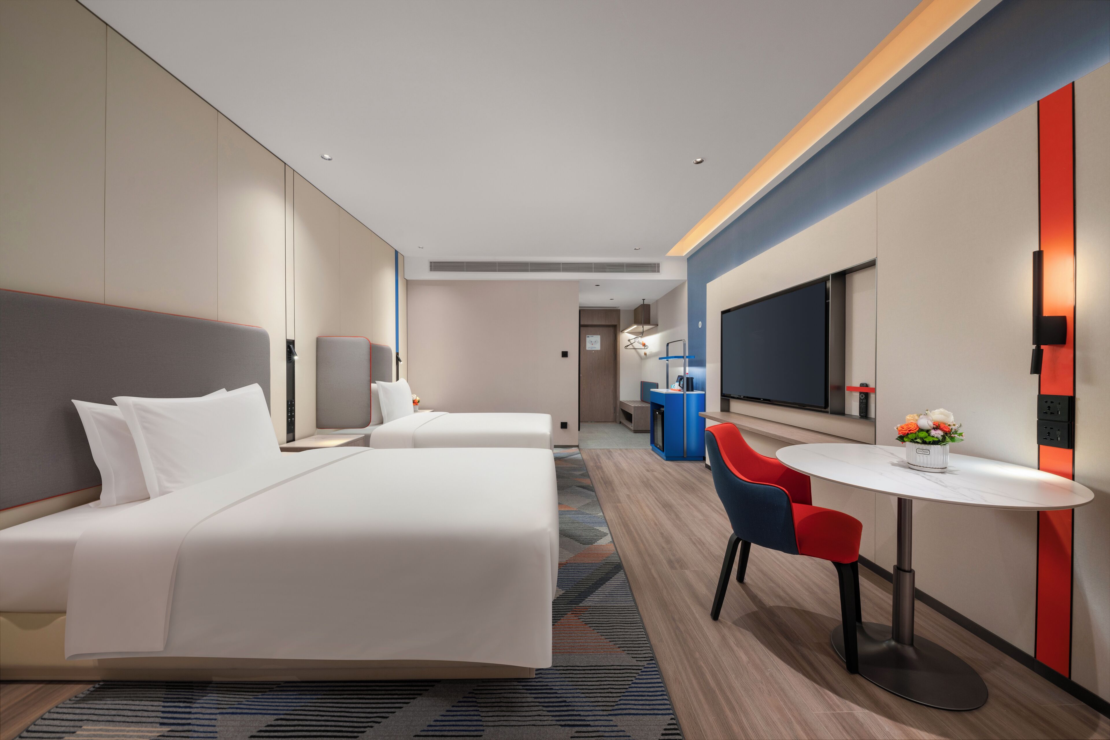 Quarto de hotel moderno com duas camas, uma mesa redonda pequena com cadeiras, televisão e escrivaninha com computador.