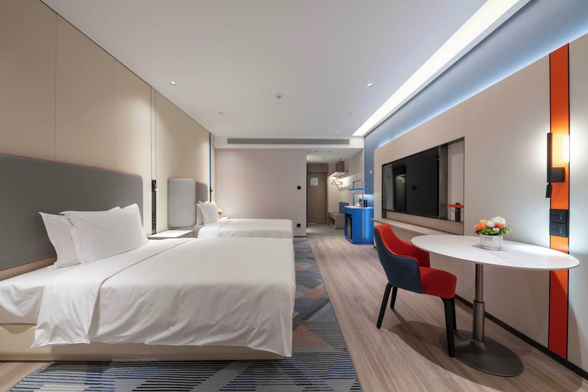 Quarto de hotel com duas camas, uma mesinha redonda com cadeiras, televisão e banheiro ao fundo.