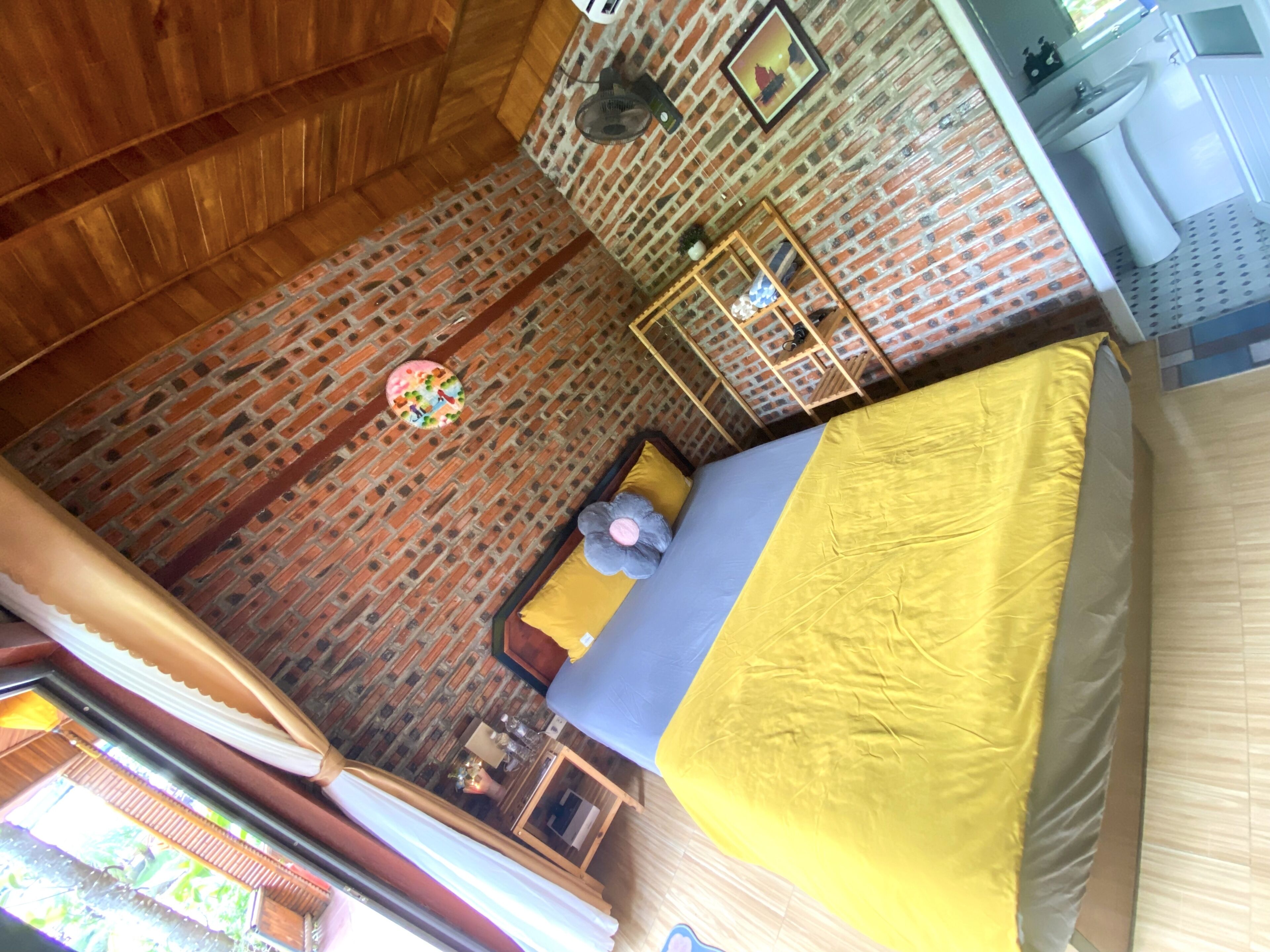 Quarto com parede de tijolos, cama com colcha amarela, mesa redonda pequena com toalha de mesa colorida e prateleira de madeira com diversos objetos.