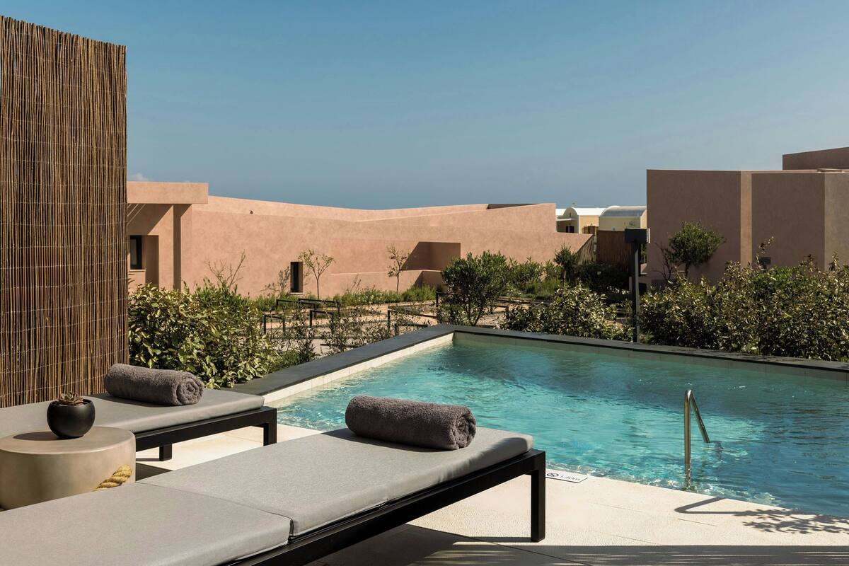 Uma área à beira da piscina com espreguiçadeiras e uma mesa pequena, cercada por vegetação e edifícios.