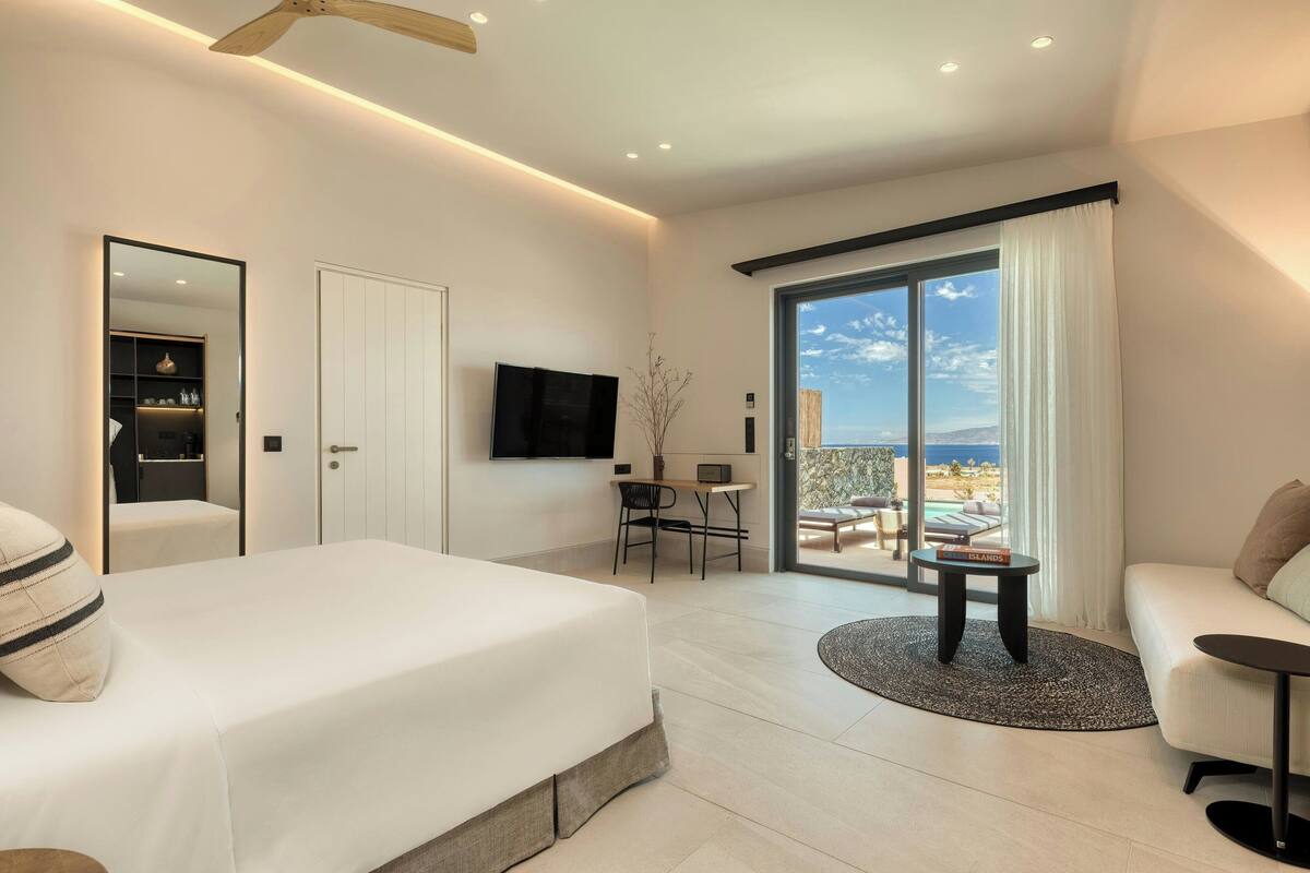 Quarto de hotel moderno com uma cama grande, televisão, escrivaninha com cadeira, sofá e vista para o mar.
