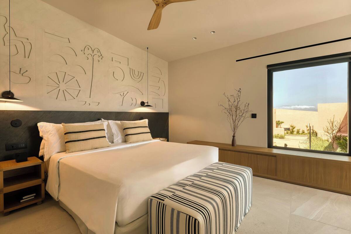 Quarto moderno com uma cama grande, mesa de cabeceira de madeira, uma mesinha, janela com vista e arte abstrata na parede.