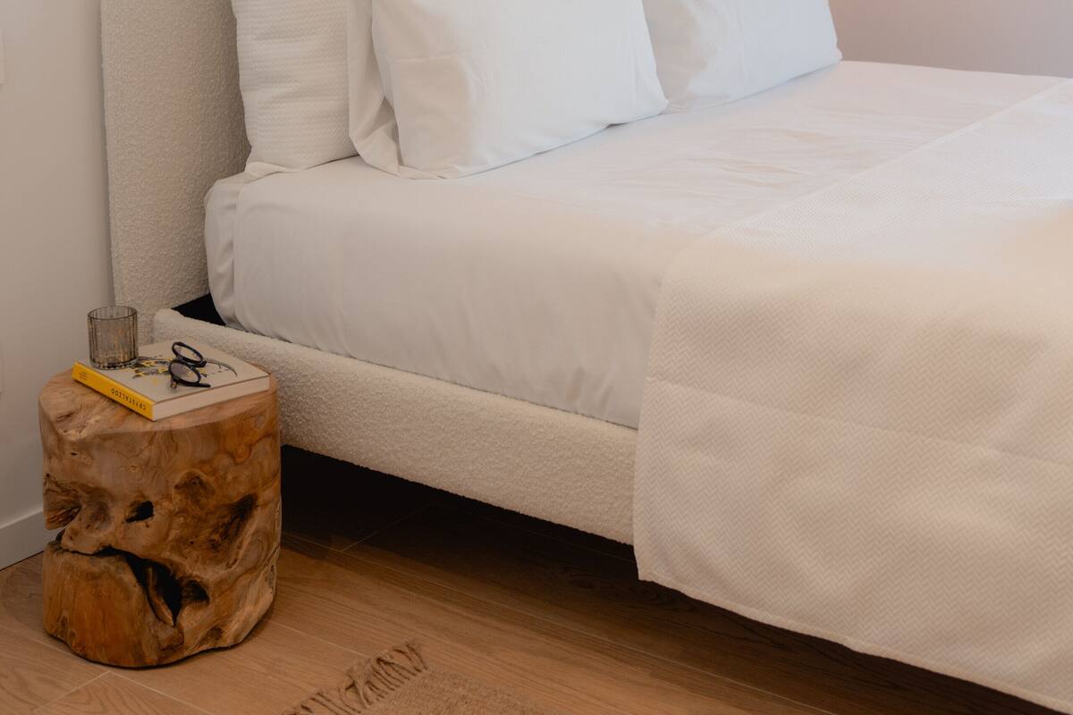 Uma cama bem arrumada com roupa de cama branca, uma mesa de cabeceira redonda de madeira com um livro e óculos, e um lustre suspenso.