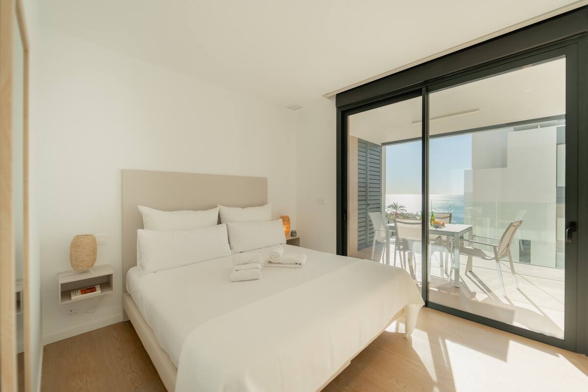 Quarto moderno com uma cama grande, uma mesinha de cabeceira e vista para uma varanda com móveis de exterior.