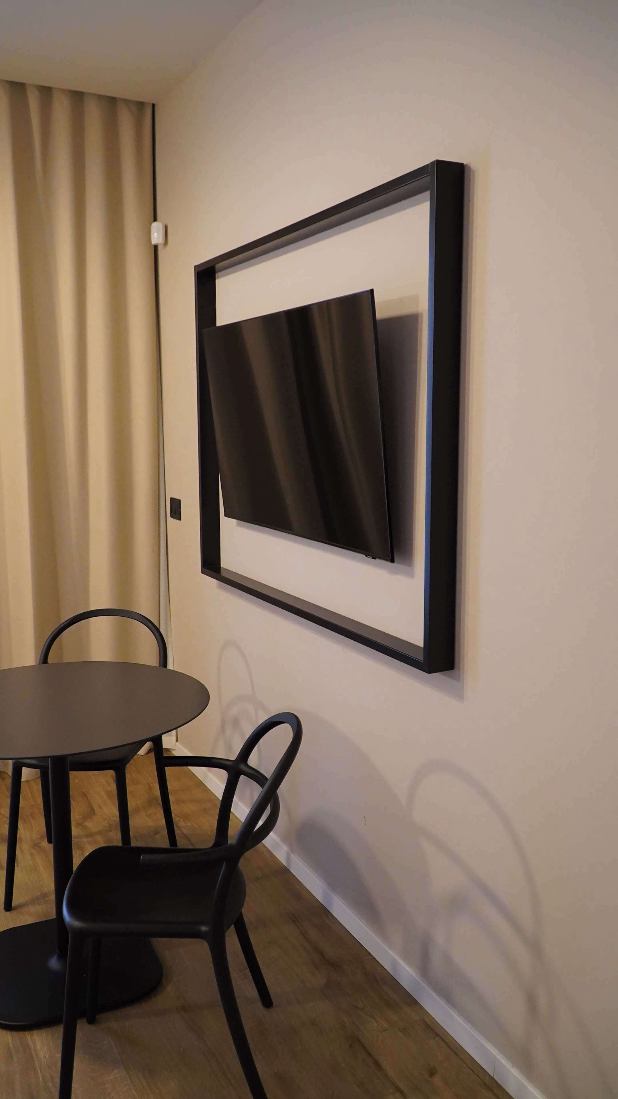 Quarto com mesa de jantar redonda e duas cadeiras, televisão de tela plana na parede e janela com cortinas bege.