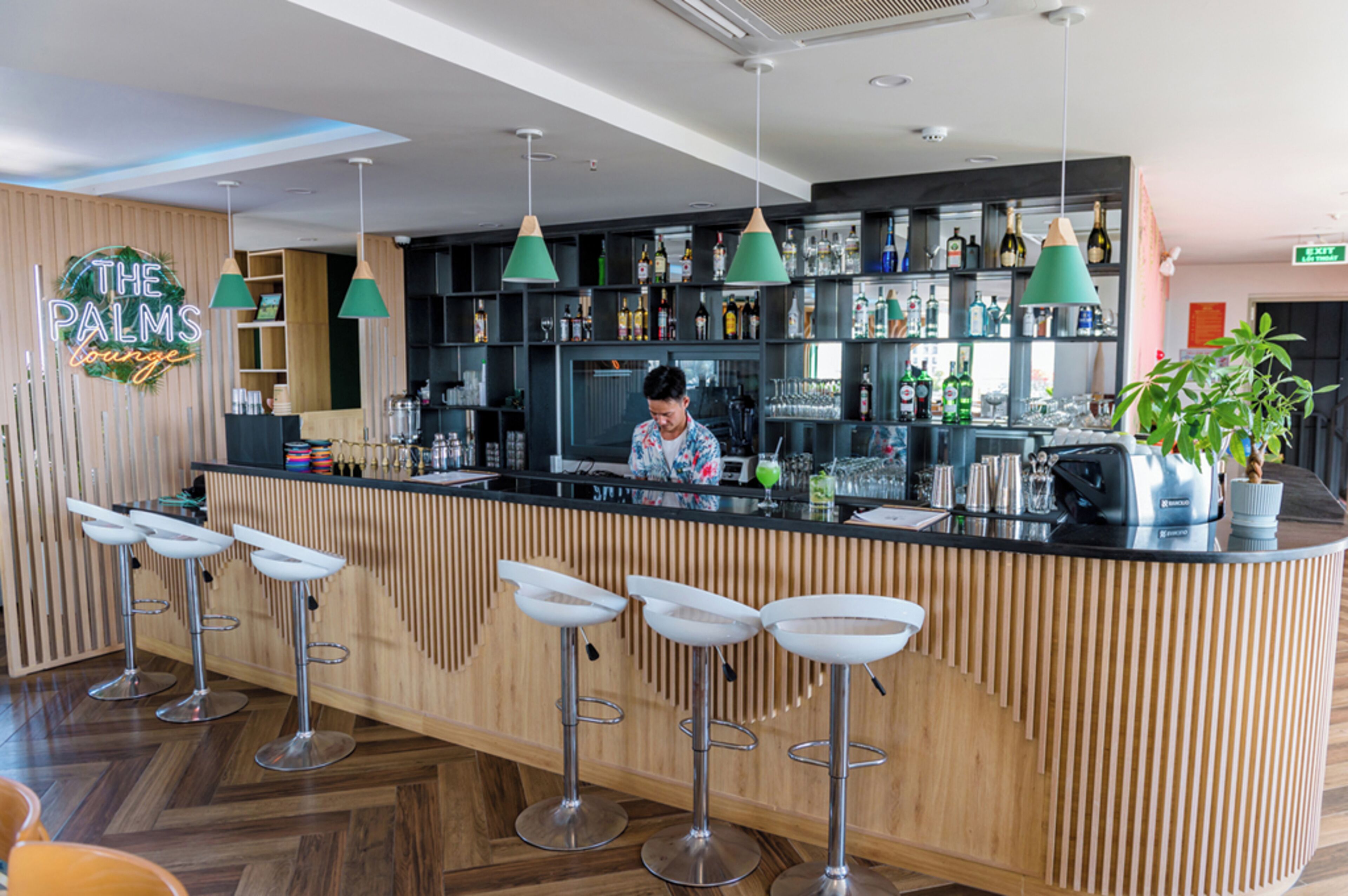 Uma área de bar com balcão de madeira, vários bancos, luminárias pendentes verdes e prateleiras com garrafas.