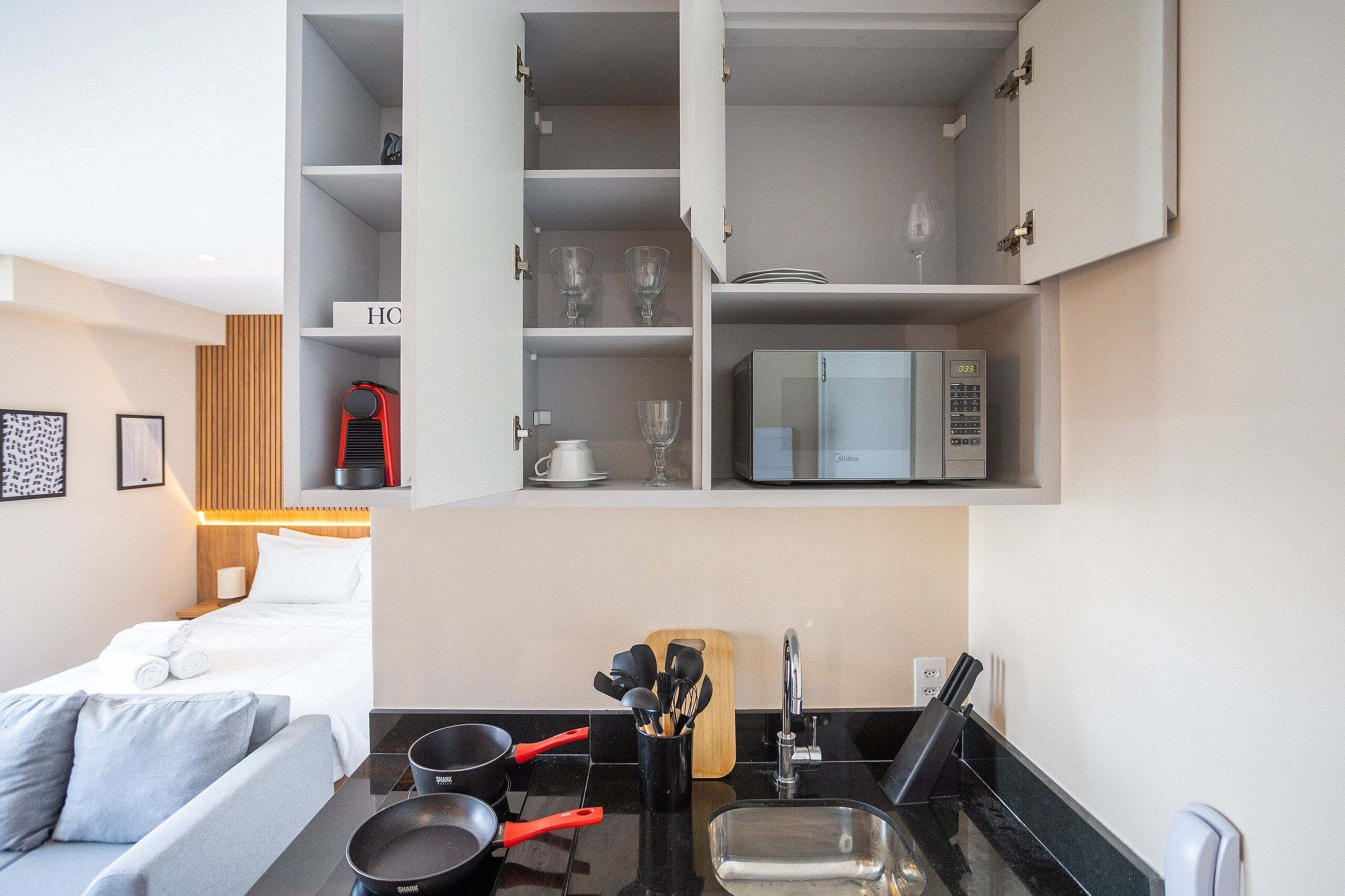 Quarto de hotel pequeno com cozinha compacta, cama com roupa de cama branca, sofá cinzento e painel de parede em madeira.
