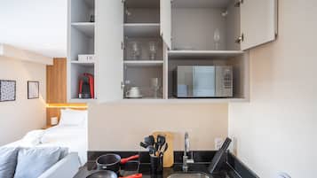 Quarto de hotel pequeno com cozinha compacta, cama com roupa de cama branca, sofá cinzento e painel de parede em madeira.