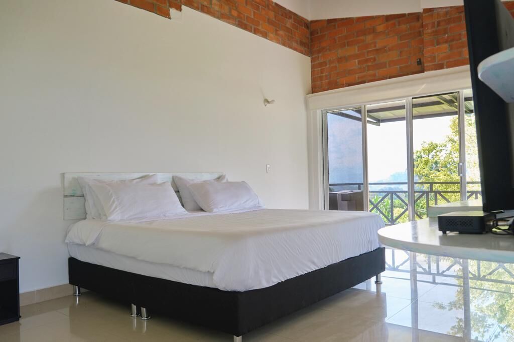 Quarto com cama grande, roupa de cama branca, parede de tijolos e varanda com mesa e cadeira.
