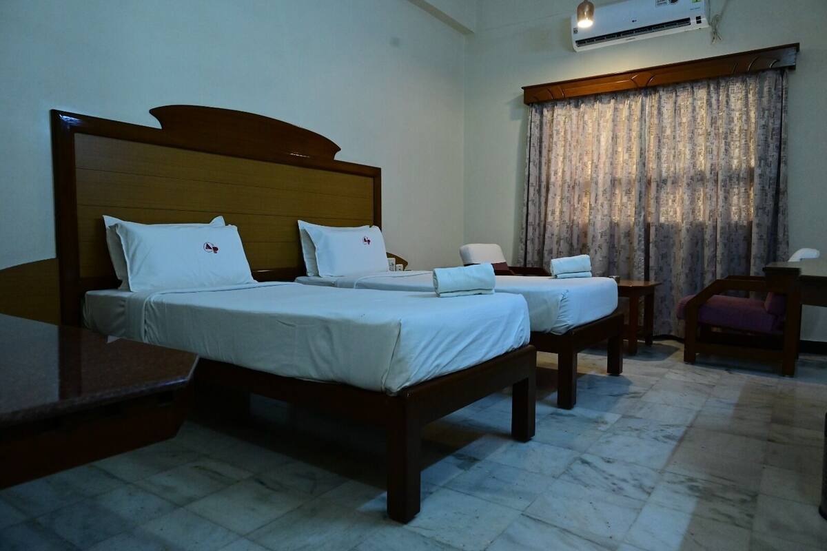 Quarto de hotel com duas camas de solteiro, cabeceira de madeira, uma mesinha, uma cadeira e janela com cortinas.