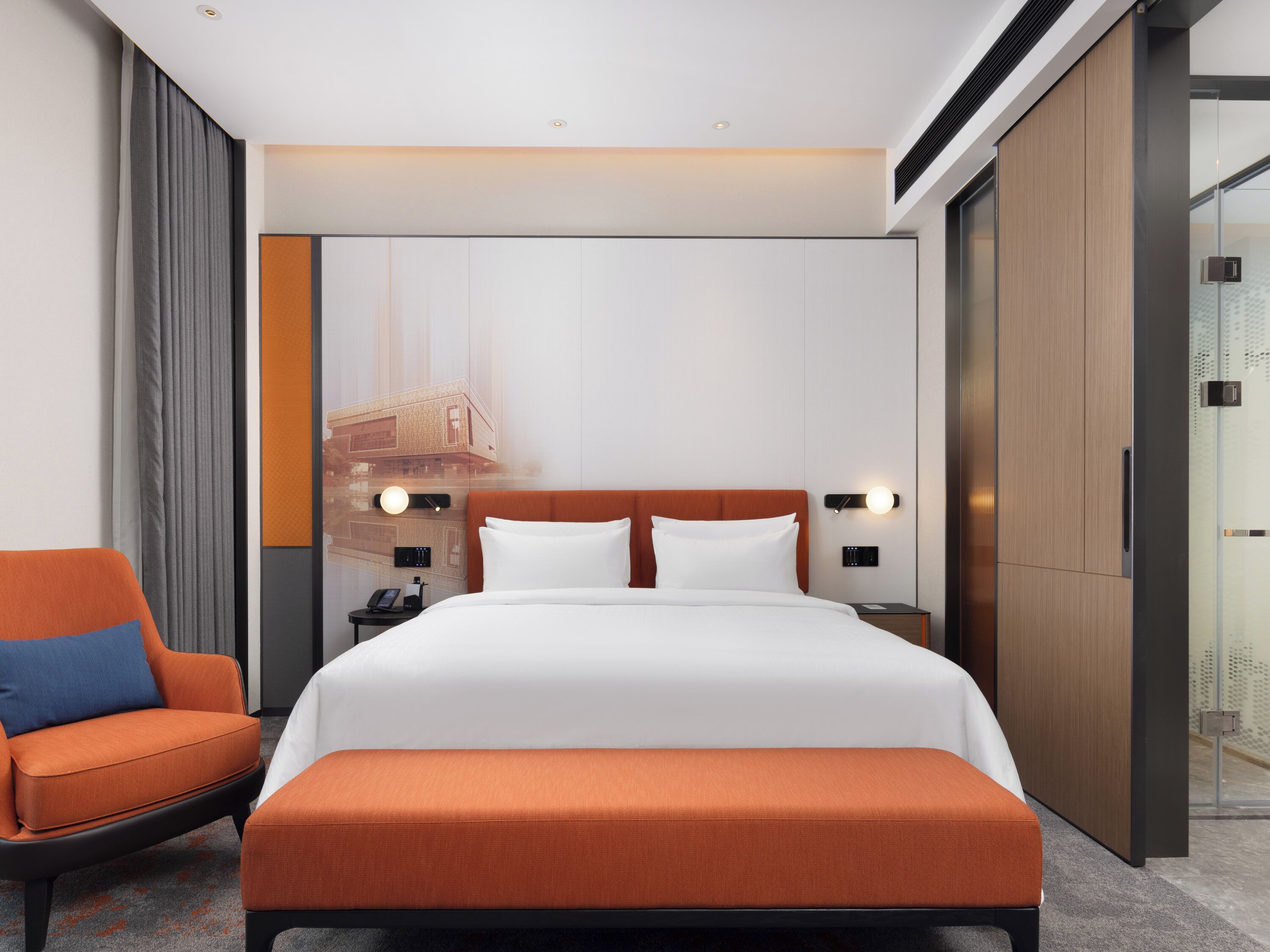 Quarto de hotel moderno com uma cama grande, área de estar com uma poltrona laranja e divisória de vidro.