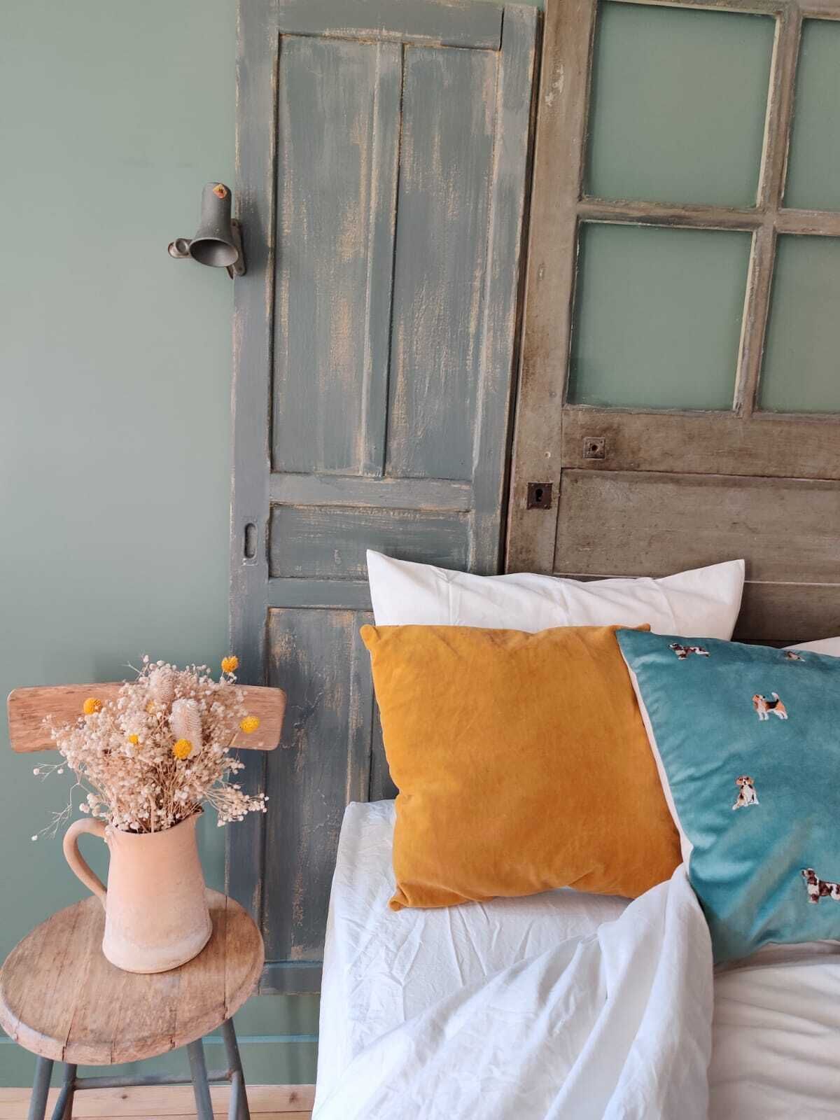 Quarto com porta de madeira rústica, um pequeno banquinho de madeira e um vaso de flores secas, e uma cama com roupa de cama branca e almofadas decorativas.