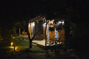 Um pequeno edifício rústico com fachada de tijolos e varanda de madeira. Há várias cadeiras e uma mesa na varanda, e alguns galos podem ser vistos em primeiro plano.