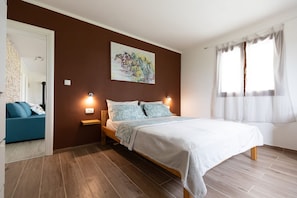Um quarto com cama, mesa de cabeceira, pintura na parede, janela com cortinas e um sofá azul no quarto vizinho.