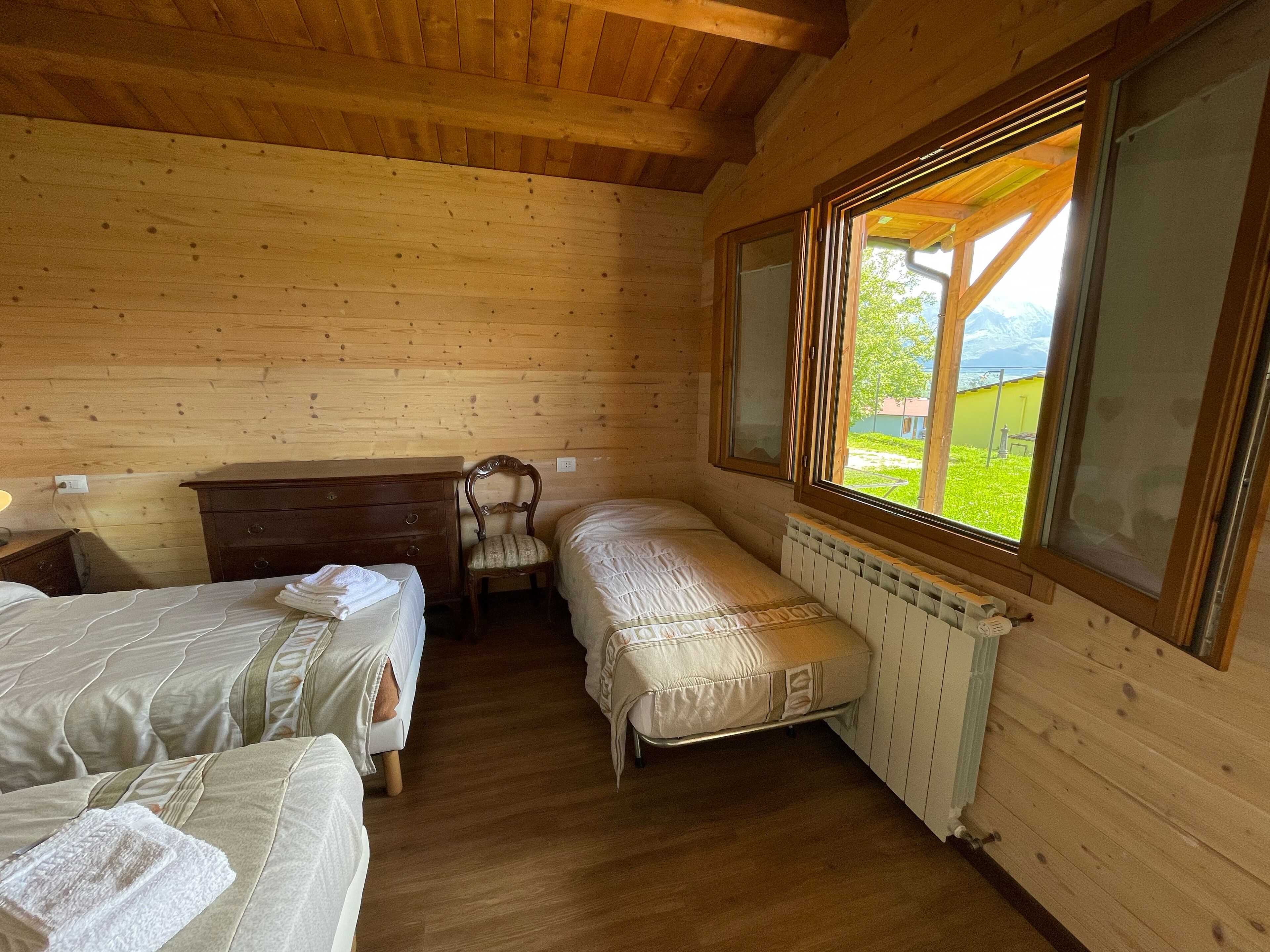 Quarto de cabana de madeira com duas camas, uma cômoda de madeira, uma cadeira e uma janela com vista para o verde.