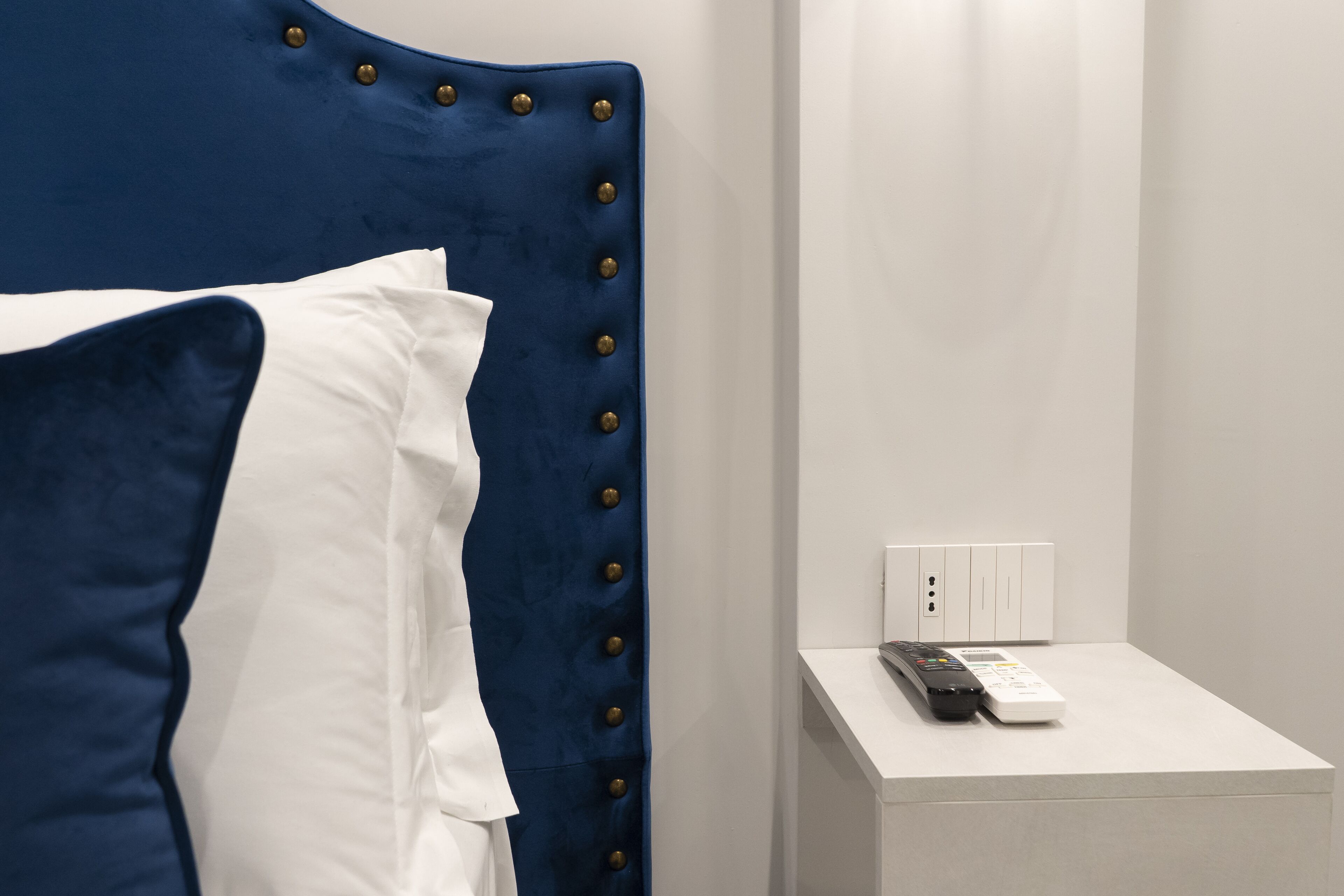 Uma cama com cabeceira azul e almofadas brancas, uma mesa de cabeceira com controle remoto e uma parede branca com tomada.