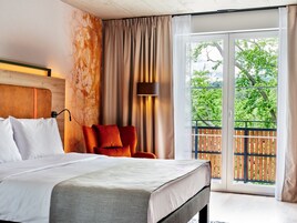 Quarto de hotel com cama, uma cadeira, uma janela grande e vista para o verde lá fora.