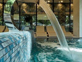 Piscina interior moderna com um característico elemento aquático curvo, espreguiçadeiras e vista para o verde através de grandes janelas.