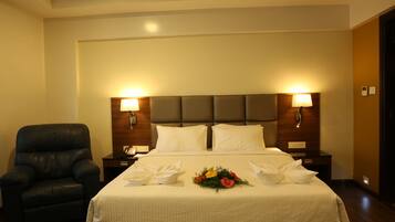 Quarto de hotel com uma cama grande, duas luminárias de cabeceira, uma poltrona de couro e um arranjo floral sobre a cama.