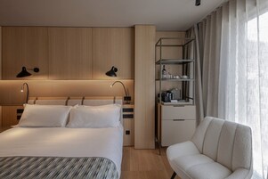 Quarto de hotel moderno com uma cama grande, abajures, uma escrivaninha com cadeira e uma janela com cortinas.