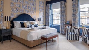 Quarto com cama grande, mesa de cabeceira, um banquinho, poltronas com listras e vista para o exterior.