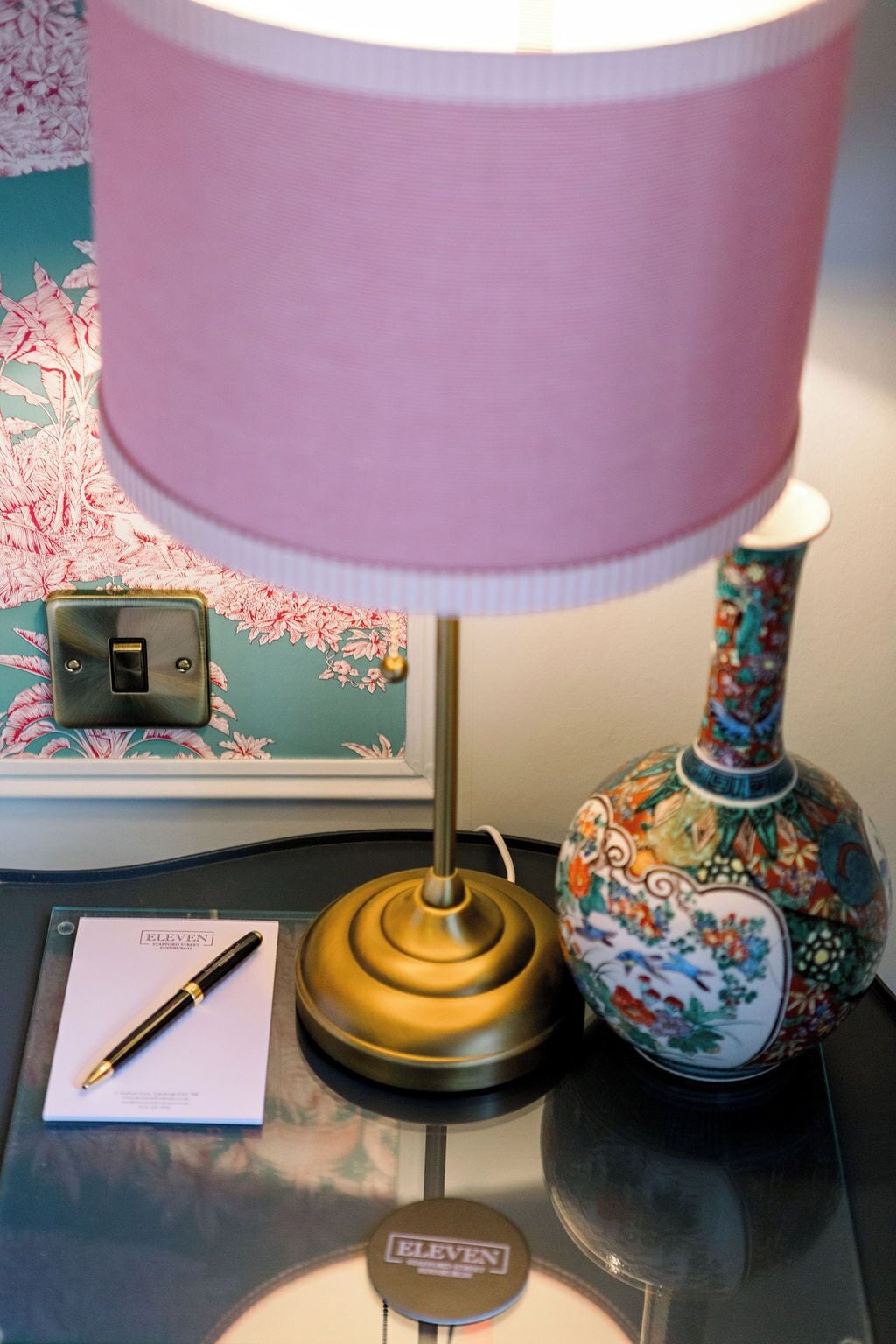 Uma luminária com cúpula rosa, um vaso decorativo, uma caneta e um bloco de notas sobre uma mesa.