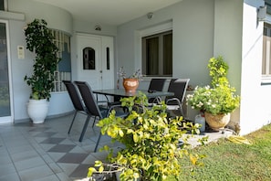 Um pátio externo com mesa e cadeiras, rodeado por plantas em vasos e uma grande janela.