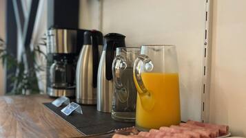Uma mesa de café da manhã com copo de suco de laranja, cafeteira e um prato de presunto e queijo.