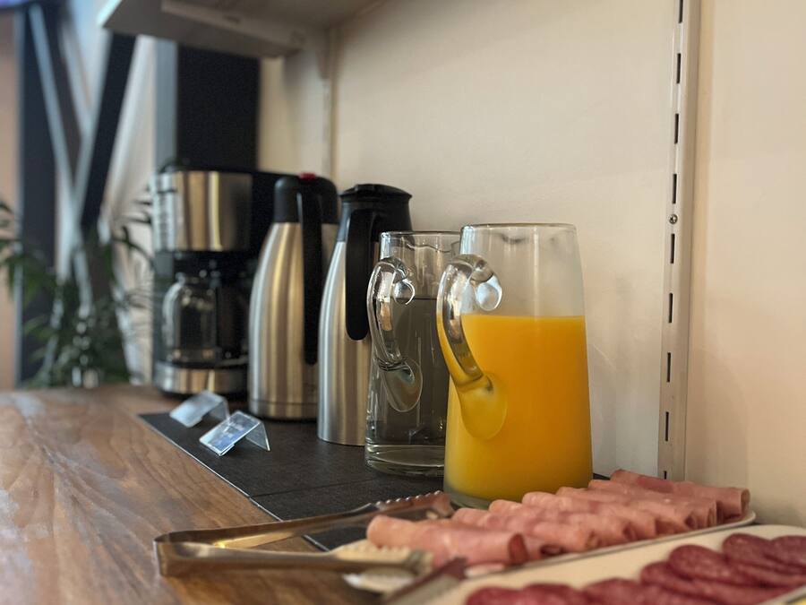 Uma mesa de café da manhã com copo de suco de laranja, cafeteira e um prato de presunto e queijo.