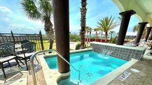 Piscina com borda azulejada, cercada por palmeiras e um espaço de terraço com mobiliário de exterior.