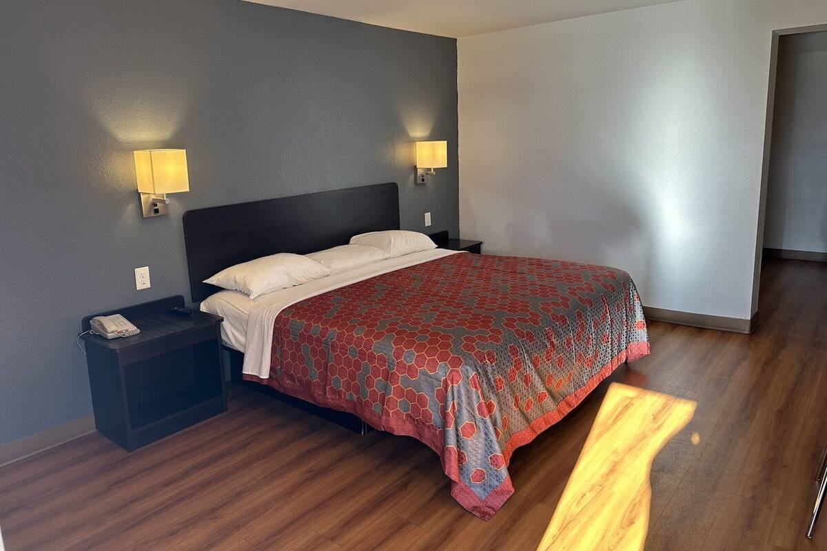 Cama bem arrumada com colcha estampada, duas luminárias de cabeceira, mesa de cabeceira com telefone e piso de madeira.