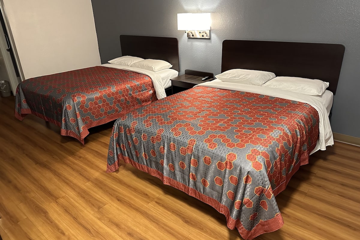 Duas camas de solteiro com colchas vermelhas e cinzentas estampadas, um criado-mudo com abajur e piso de madeira.