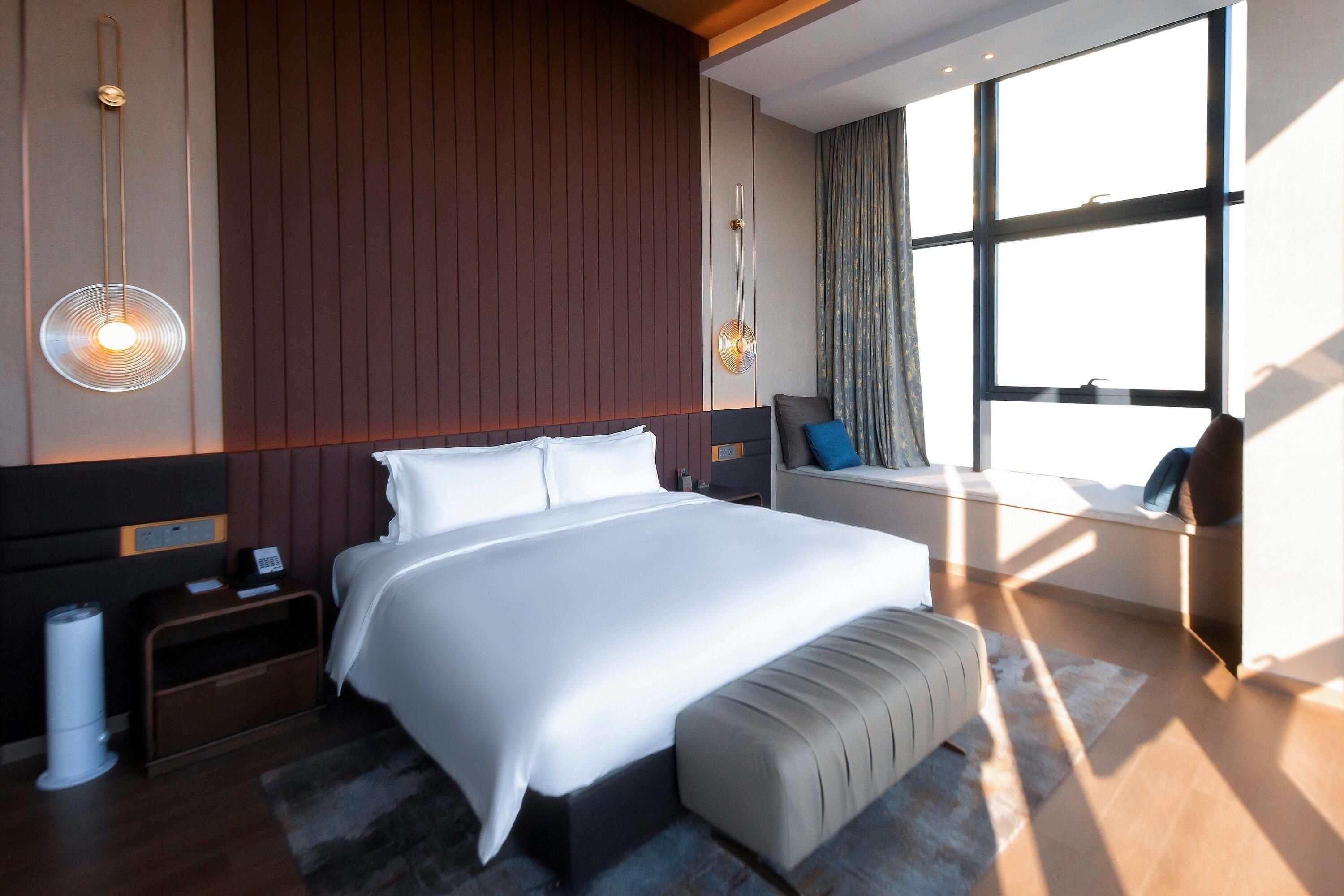 Quarto de hotel moderno com uma cama grande, mesas de cabeceira, abajures, uma janela com cortinas e vista para o exterior.