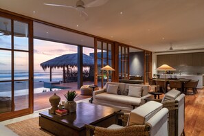 Uma sala de estar ampla com uma grande janela para a praia, um espaço de estar aconchegante e uma cozinha moderna ao fundo.