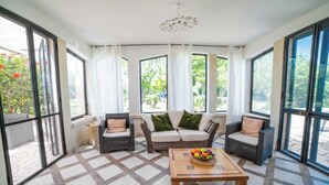 Um quarto espaçoso com grandes janelas, um sofá central, duas poltronas, uma mesa de centro com frutas e um piso estampado.