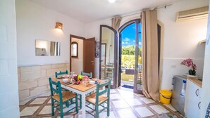Uma sala de jantar com mesa e cadeiras de madeira, uma cozinha com geladeira, e vista através de uma porta aberta.
