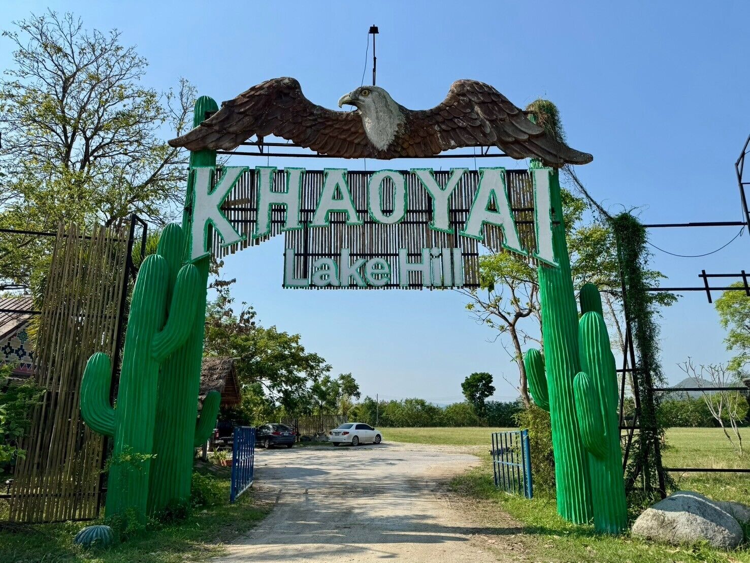 Um letreiro com uma águia e as palavras "KHAOYAI LAKEH HI" em um cenário rústico com cactos e uma estrada de terra.