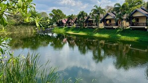 Uma fileira de acomodações em estilo bangalô, com varandas, um lago calmo em primeiro plano e vegetação exuberante ao redor da propriedade.