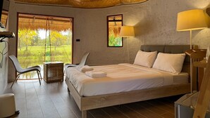 Quarto com cama, duas luminárias de cabeceira, uma cadeira, uma mesinha e uma janela com vista para o verde.