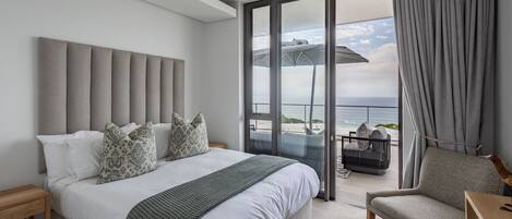 Quarto de hotel moderno com uma cama grande, varanda e vista para o mar, e uma cadeira.