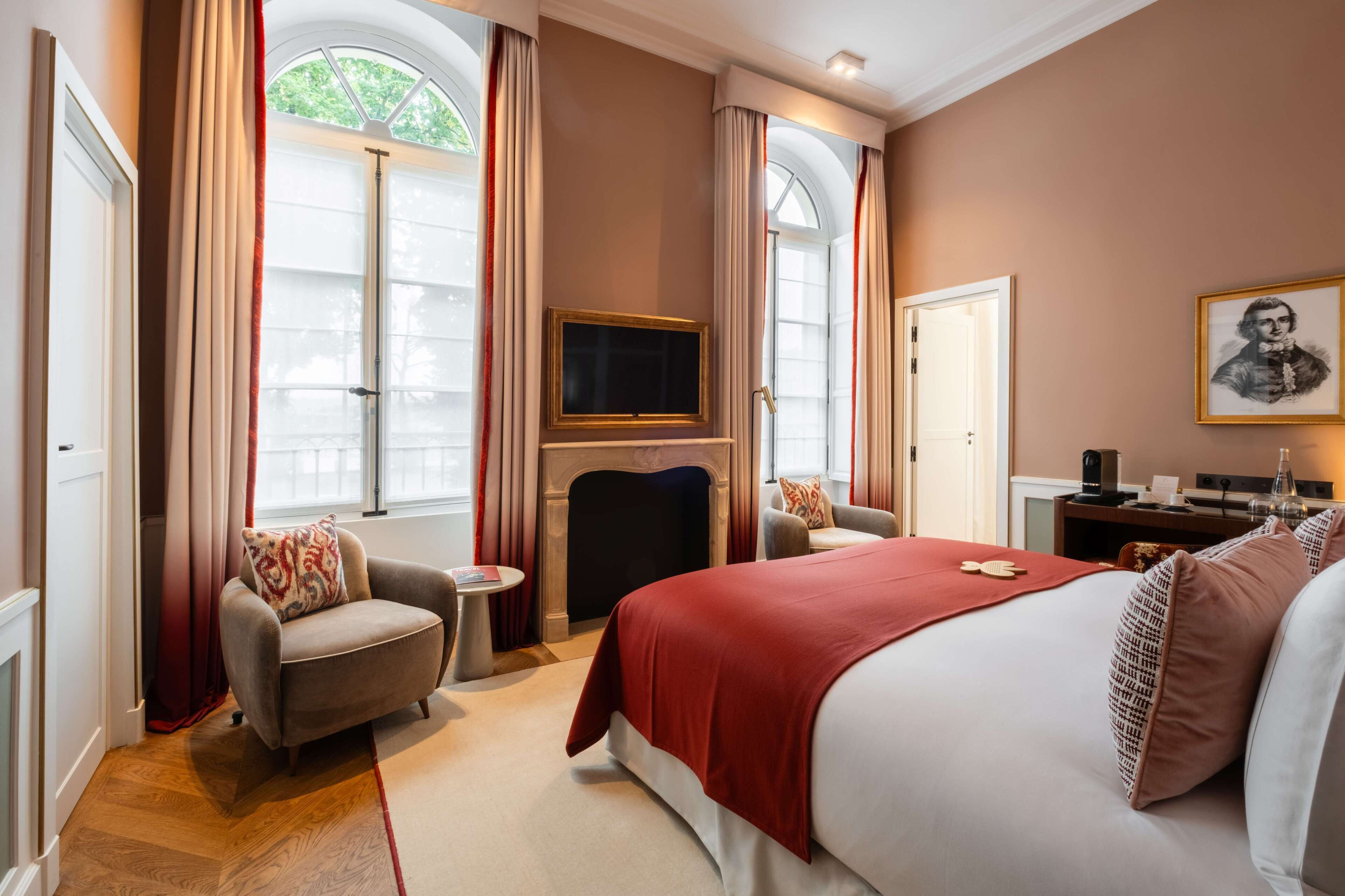 Quarto de hotel com uma cama grande, duas poltronas, uma mesa pequena, lareira, televisão e um retrato na parede.