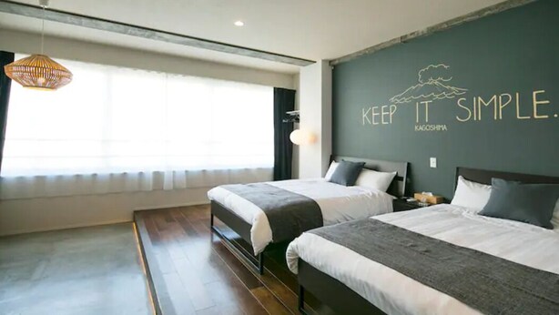 Quarto de hotel moderno com duas camas, uma janela grande e uma parede com uma frase motivacional.