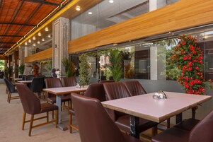 Um restaurante moderno com mesas de madeira e assentos em couro, amplas janelas com plantas e teto alto.