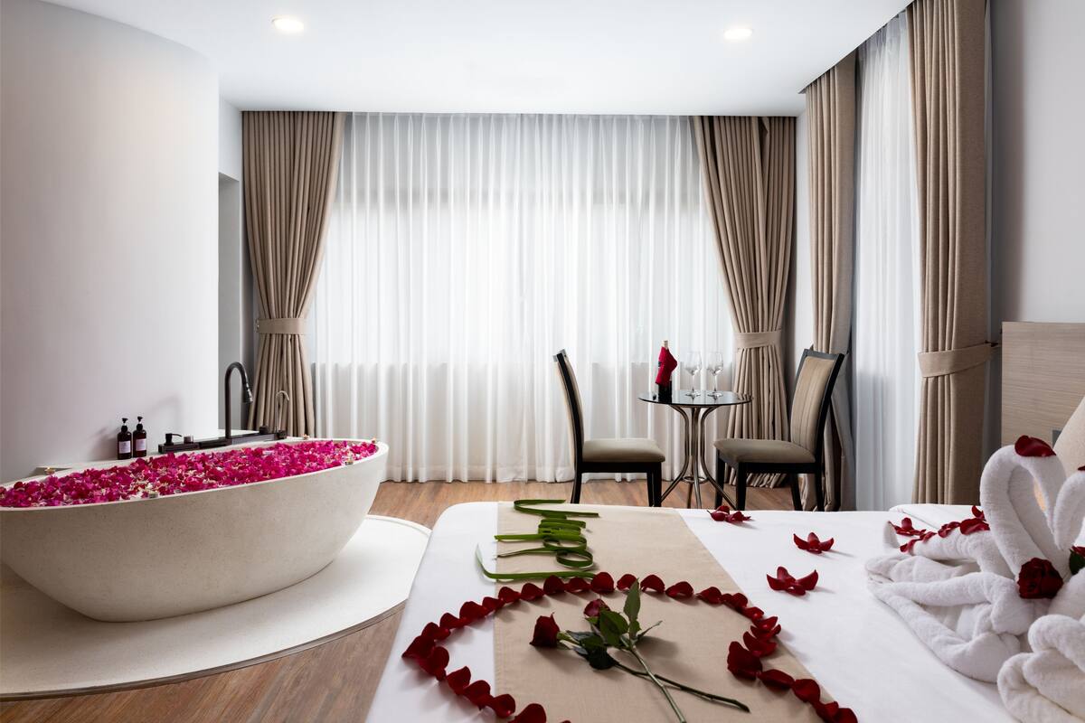 Quarto de hotel moderno com uma banheira grande cheia de pétalas de rosa, uma cama bem arrumada com pétalas e um buquê, e uma mesa com um vaso de flores.