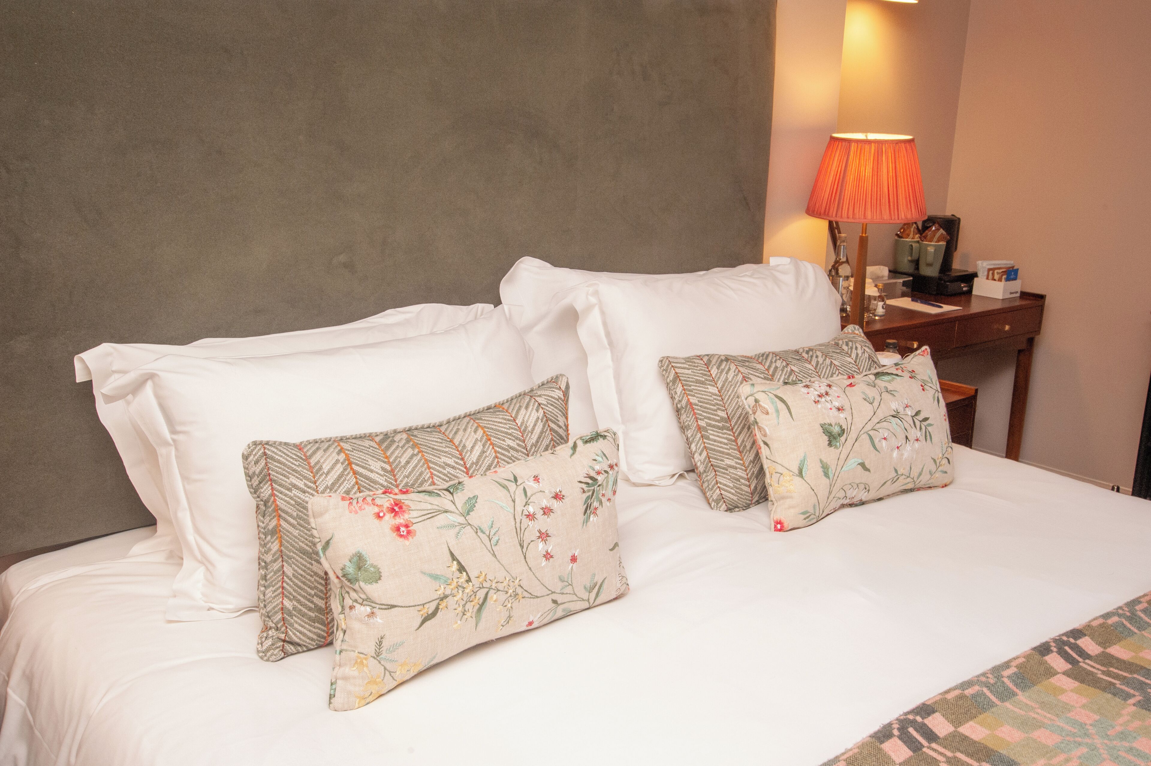 Uma cama bem arrumada com roupa de cama branca e almofadas estampadas com flores, uma mesa de cabeceira com abajur e uma parede com acabamento texturizado.