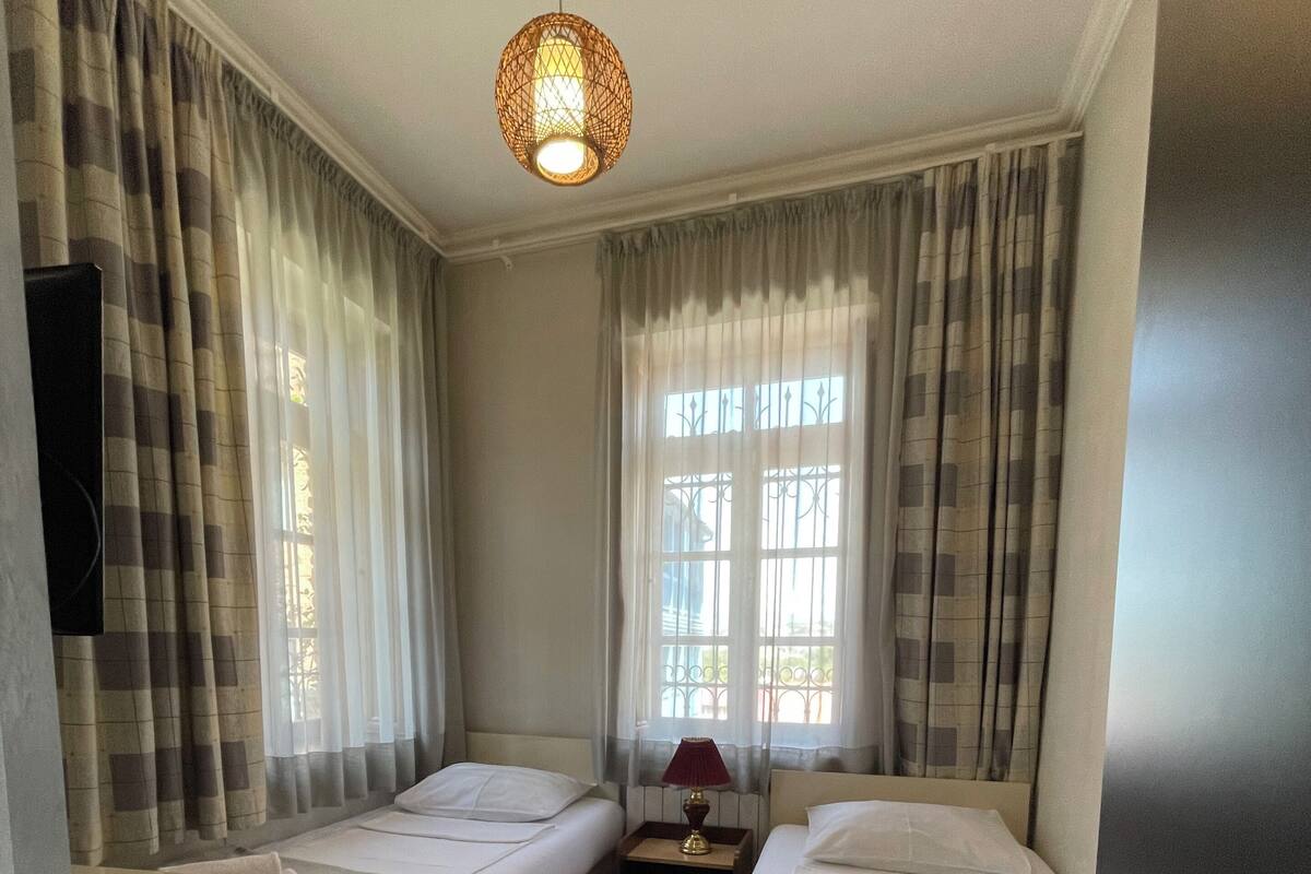 Quarto de hotel com duas camas de solteiro, piso de madeira, mesa de cabeceira pequena com luminária e janela com cortinas.
