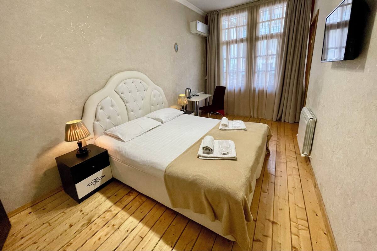 Quarto de hotel com uma cama grande, mesa de cabeceira com abajur, uma cadeira, pequena mesa com chaleira e uma TV fixada na parede.